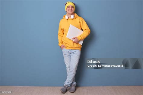 비니 모자와 캐주얼한 노란색 후드티를 입고 파란 벽에 포즈를 취하고 카메라를 보고 닫힌 노트북을 들고 작업을 마치고 기뻐하는