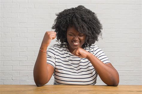 젊은 아프리카계 미국인 여자 집에서 매우 행복 하 고 흥분 무기와 우승자 제스처를 하 고 테이블에 앉아 웃 고 성공을 위한 비명 축 하 개념입니다 T 셔츠에 대한 스톡 사진