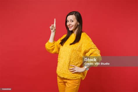 노란색 모피 스웨터에 웃는 예쁜 젊은 여자의 초상화 스튜디오에서 밝은 빨간색 벽 배경에 고립 된 인덱스 손가락을 가리키는 사람들은 진심으로 감정 라이프 스타일 개념입니다