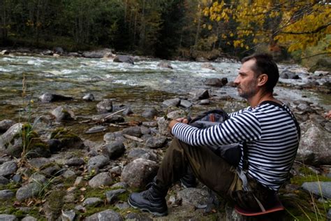 Фотография на тему Мужчина с бородой в жилете сидит на берегу чистой горной реки в окружении