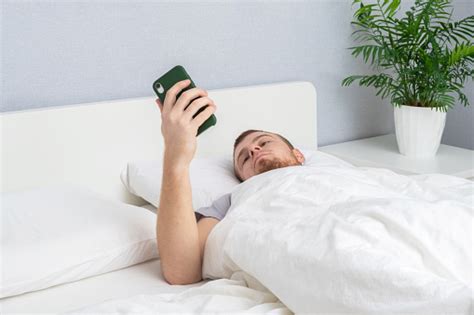 수염을 기른 남자는 집 침대에서 전화를 사용합니다 침대에서 일어나지 않고 물건을 쇼핑하십시오 가제트에 대한 사람들의 의존 개념