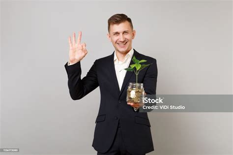 회색 배경에 고립 된 녹색 식물 새싹과 유리 항아리에 더미 금화를 들고 확인 제스처를 보여주는 잘생긴 젊은 비즈니스 남자 업적