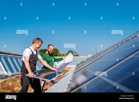 Craftsman Installing Solar Panel On The Roof Of A Company Building With Colleague Holding Plan