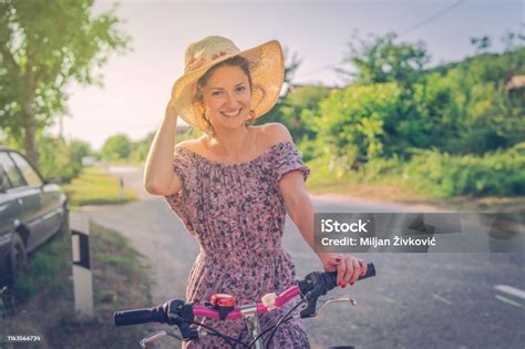 야외 오픈 로드 아스팔트에서 드레스와 모자를 입고 여름 화창한 날에 자전거를 밀어 가는 핸들 바 그립을 들고있는 젊은 여성