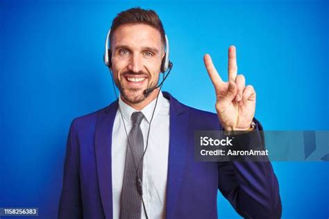 파란색 고립 된 배경 위에 콜 센터 헤드셋을 착용한 젊은 잘생긴 연산자 남자가 승리의 사인을하는 카메라에서 행복한 얼굴 윙크와 미소 2번 경영자에 대한 스톡 사진 및 기타