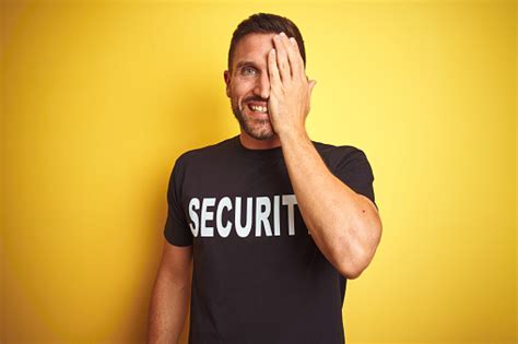 한 눈을 손으로 덮고 얼굴에 자신감 있는 미소와 깜짝 감정을 감싸는 노란색 고립된 배경 위에 보안 유니폼을 입은 젊은 보호대