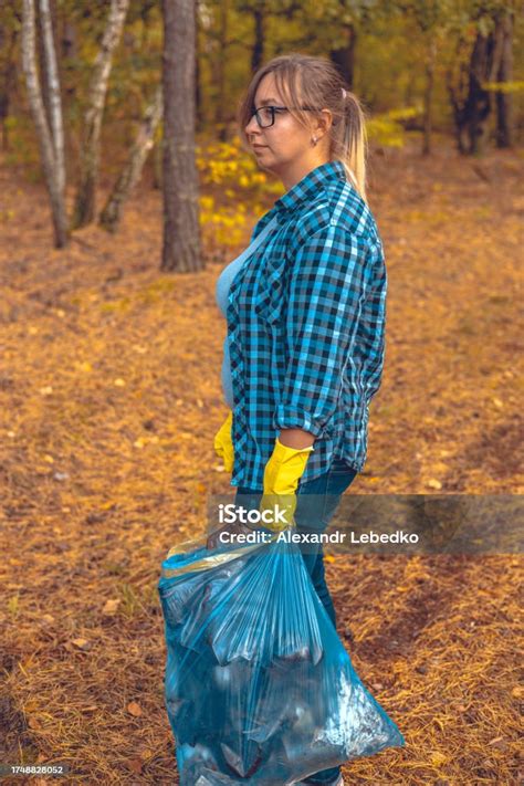 숲에서 쓰레기를 치우고 피곤한 여성 자원 봉사자가 쓰레기 봉투를 손에 들고 서서 슬프게 옆을 바라본다 한 여성이 쓰레기로 인한 음식과 오염에 대해 걱정합니다 가방에 대한