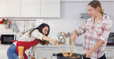 귀여운 즐거운 커플이 함께 요리하고 식사에 향신료를 더하고 부엌에서 함께 웃고 시간을 보내고 앞치마를 입은 레즈비언 커플 프라이팬이 있는 나무 테이블 뒤에서 함께 웃고
