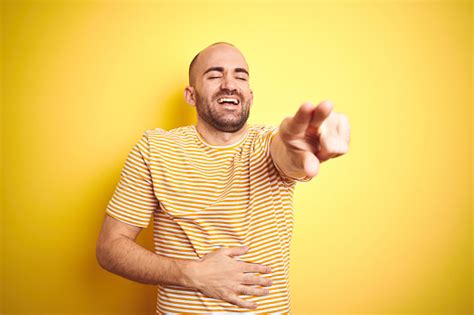 노란 고립 된 배경 위에 캐주얼 스트라이프 티셔츠를 입고 수염을 가진 젊은 대머리 남자는 몸을 손으로 카메라에 손가락을 가리키고