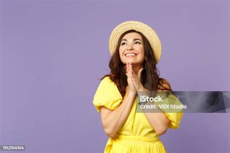 노란 드레스에 웃는 젊은 여자 여름 모자를 찾고 접힌 손을 만드는 소원 파스텔 바이올렛 벽 배경에 고립 된 기도 사람들은 진심으로 감정 라이프 스타일 개념입니다 복사 공간을