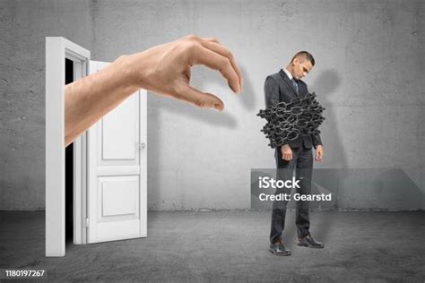 슬픈 작은 사업가의 전체 길이 측면보기 그의 몸 주위에 체인과 거대한 손이 그의 뒤에 문에서 나오는 서 개념에 대한 스톡 사진