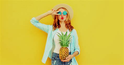 파인애플이 빨간 입술을 불고 있는 아름다운 여성의 여름 초상화가 밀짚 모자를 쓰고 선글라스를 쓰고 화려한 노란색 배경을 가진