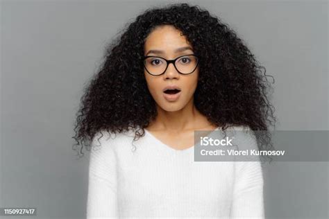 어두운 곱슬 머리와 안경에 충격 아름 다운 여자의 솔직 한 샷 놀랍게도에서 떨어졌다 턱을 유지 놀라운 무언가를 발견 한 후 무감각 회색 배경 위에 고립 Omg에 대한 스톡