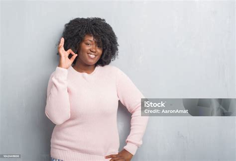 젊은 아프리카계 미국인 플러스 크기 여자 회색 그런 지 벽 입고 겨울 스웨터 웃 고 긍정적인 손과 손가락으로 Ok 사인을 하 고 이상 성공적인 식입니다 가을에 대한 스톡