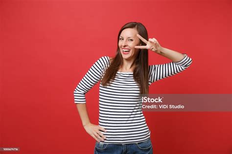 캐주얼 스트라이프 옷을 입고 서 있는 밝은 빨간 벽 배경에 고립 승리 기호를 보여주는 웃는 예쁜 젊은 여자의 초상화 사람들이