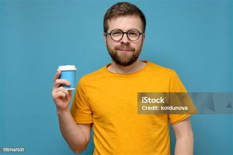 둥근 안경과 노란색 티셔츠를 입은 침착하고 차분한 남자가 일회용 종이 컵을 손에 들고 파란색 배경에 있습니다 귀여운에 대한 스톡 사진 및 기타 이미지 Istock