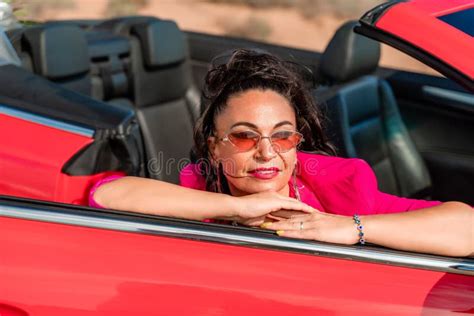 A Woman In A Pink Jacket Is Sitting In A Red Convertible She Is