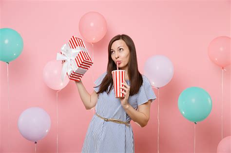 블루 드레스 선물 빨간색 상자를 들고 찾고 예쁜 여자의 초상화 선물 마시는 소 다 또는 콜라 파스텔 핑크 배경에 플라스틱 컵에서 화려한 공기 풍선 생일 휴가 파티 14에