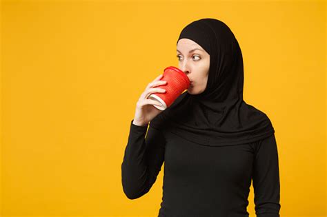 젊 피곤한 슬픈 화가 아라비아 무슬림 여자에 히 잡 블랙 의류 노란 벽 배경 스튜디오 초상화에 고립 된 커피의 종이 컵을 개최
