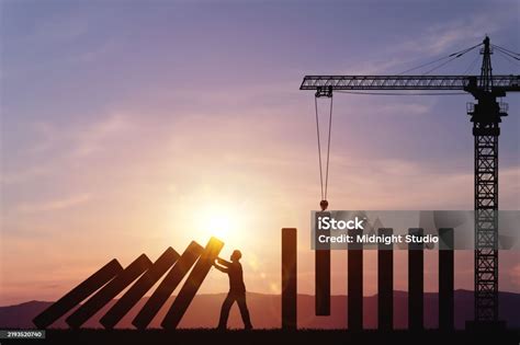 도미노를 멈추기 위해 직사각형 블록을 아래로 밀어내는 남자의 실루엣과 함께 밝은 하늘 일몰을 배경으로 직사각형 블록을 쌓는 크레인 개념에 대한 스톡 사진 및 기타 이미지
