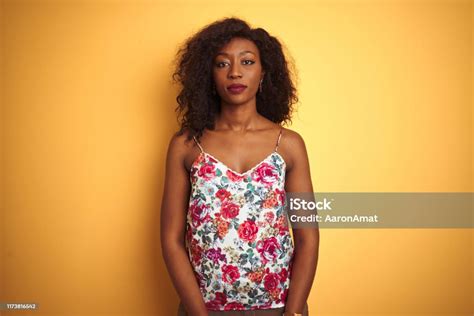 고립된 노란색 배경 위에 꽃무늬 여름 티셔츠를 입은 아프리카계 미국인 여성이 얼굴에 진지한 표정을 짓고 있습니다 카메라를 보는 간단하고 자연스러운 T 셔츠에 대한 스톡 사진