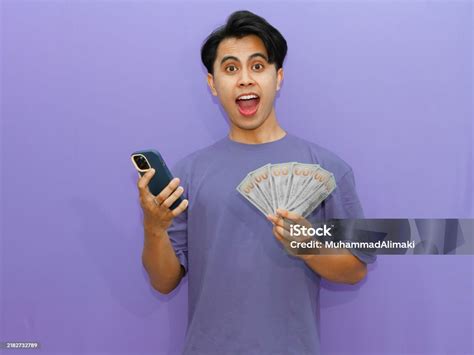 놀란 표정을 가진 젊은 아시아 남자 돈을 들고 휴대 전화를 사용 보라색 배경에 고립되어 있습니다 광고 및 판촉용으로 적합합니다 Brand Name Smart Phone에