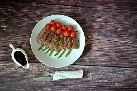 방울 토마토와 삶은 아스파라거스를 곁들인 쇠고기 스테이크를 나무 테이블에 조각으로 자른 접시 소스가 담긴 컵과 냅킨이 달린 칼 옆 고기에 대한 스톡 사진 및 기타 이미지