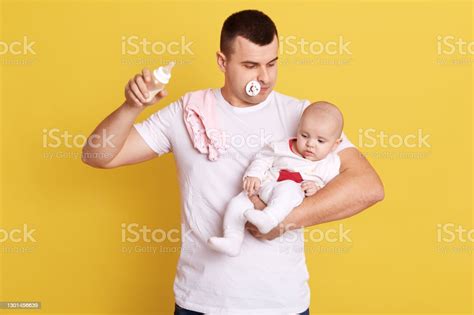 어린 아버지는 아기를 안고 우유로 아이를 먹이려고 하고 아이의 젖꼭지를 손에 들고 포즈를 취하고 아무렇지 않게 입고 있는 남자가 노란 배경위에 고립된 수유병 아빠 아기를