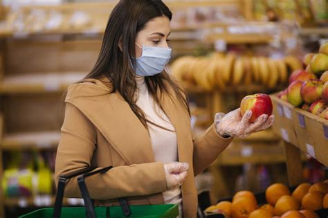 보호 장갑과 얼굴 마스크를 착용한 젊은 여성은 아름다운 신선한 사과를 손에 들고 있습니다 음식과 함께 서서 음식을 선택하는 음식 바구니와 아름다운 어린 소녀 검역 중 쇼핑