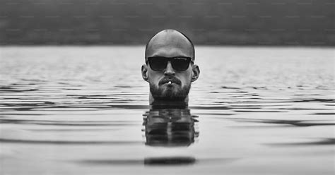 담배를 피우고 선글라스를 낀 잔인한 수염을 기른 남자가 호수에 나타납니다 비오는 안개가 낀 날 대기 순간에 호수에서 물 위에서 담배를 피우는 남자 머리 방랑벽 창조적