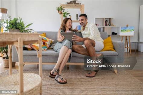 거실의 소파에 앉아 태블릿을 들고 커피 한 잔을 즐기는 와이드 샷 성인 커플은 그들을 행복하게 만드는 무언가를 보고 있다 텔레비전 산업에 대한 스톡 사진 및 기타 이미지