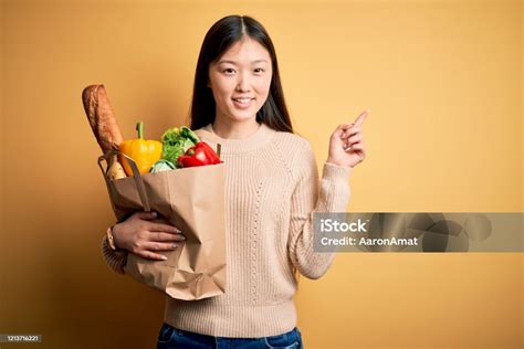 젊은 아시아 여성은 행복하고 자연스러운 표정으로 옆으로 손과 손가락으로 가리키는 얼굴에 미소와 함께 노란색 고립 된 배경을 통해 신선한 건강한 식료품의 종이 가방을 들고