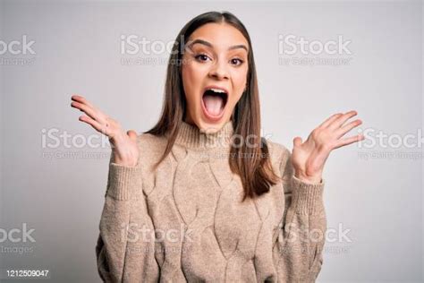 젊은 아름 다운 갈색 머리 여자 캐주얼 스웨터를 입고 흰색 배경 에 서 미친 축 하 하 고 팔을 올리고 눈을 열고 흥분 비명을