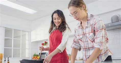 귀여운 즐거운 커플이 함께 요리하고 식사에 향신료를 더하고 부엌에서 함께 웃고 시간을 보내고 앞치마를 입은 레즈비언 커플 프라이팬이 있는 나무 테이블 뒤에서 함께 웃고