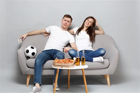 흰색 T 셔츠에 웃는 커플 여자 남자 축구 팬 응원 파이프 회색 배경에 고립 된 휴식 앉아 좋아하는 팀 지원 사람들의 감정 스포츠 가족 레저 라이프 스타일 개념입니다