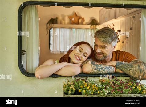 Happy Redhead Woman Smiling With Closed Eyes Near Tattooed Man In Window Of Modern Trailer Home