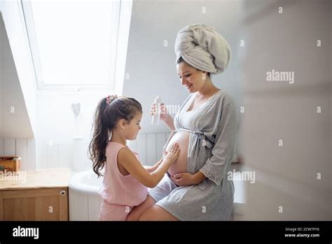 Small Girl Creaming Belly Of Pregnant Mother Indoors In Bathroom At