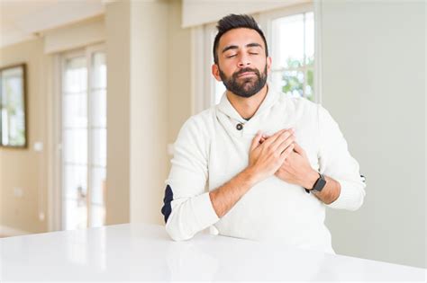 집에서 캐주얼 한 흰색 스웨터를 입은 잘 생긴 히스패닉 남자는 닫힌 눈과 얼굴에 감사 제스처와 가슴에 손을 미소 건강 개념