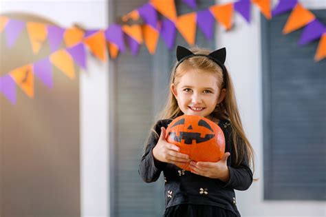 작은 고양이의 카니발 의상을 입은 웃는 소녀가 방에서 오렌지 호박과 과자를 가지고 놀고 있습니다 10월에 대한 스톡 사진 및