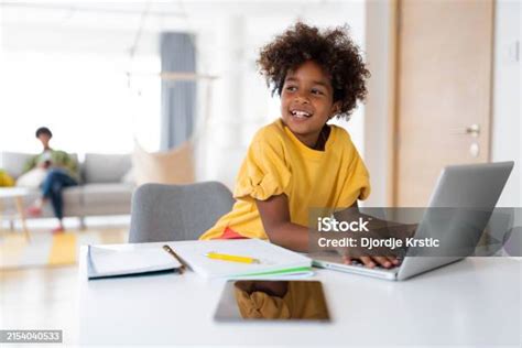 สาวแอฟริกันอเมริกันยิ้มนั่งอยู่ที่โต๊ะและใช้แล็ปท็อป แม่ของเธอนั่งอยู่บนโซฟาเป็นฉากหลั ภาพสต็อก