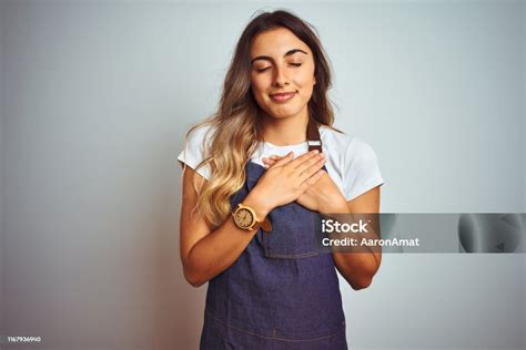 회색 고립 된 배경위에 앞치마를 입은 젊은 아름다운 여성은 닫힌 눈과 얼굴에 감사하는 제스처로 가슴에 손을 대고 미소 짓고 있습니다 건강 개념 감사에 대한 스톡 사진 및