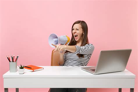 성 난 여자 검지 손가락 앉아 Pc 노트북 파스텔 핑크 배경에 고립 된 흰색 책상에서 작업을 가리키고 확성기에 비명 성과