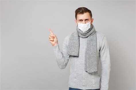 회색 스웨터에서 젊은 남자 스카프 메 마른 얼굴 마스크 포인팅 검지 손가락 옆으로 회색 벽 배경에 고립 건강 한 라이프 스타일 아픈 아픈 질병 치료 추운 계절 개념 복사