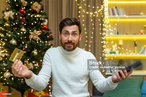 불만족스럽고 실망한 남자는 크리스마스와 새해를 맞이하여 은행 신용 카드와 스마트폰을 들고 온라인 쇼핑을 하려고 초조하게 카메라를 바라보고 있다 사기꾼에 대한 스톡 사진 및