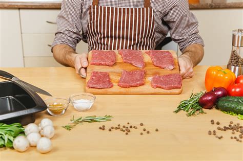 멋진 주방의 전체 빛 부엌에 돼지고기 쇠고기 또는 양고기 고기 스테이크를 준비 하는 가정에서 요리 앞치마 조각 나무 보드에 고기 야채와 함께 테이블에 앉아 있는 젊은 남자를