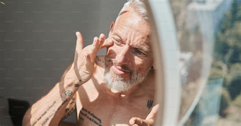 성숙한 남자는 욕실에서 거울을 보면서 얼굴에 노화 방지 크림을 바르고 있다 얼굴 피부 관리 및 위생의 개념 국내 생활 방식 문신을 한 회색 머리 백인 남성 연금 수급자