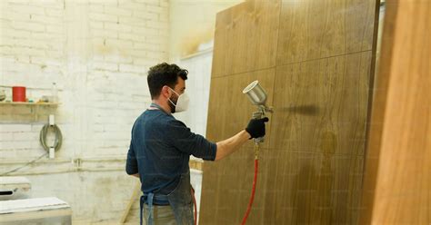 장갑을 끼고 마스크를 쓴 라틴 남성 노동자가 스프레이 건을 들고 목공소에서 갈색 나무 문을 칠하고 있다 사진 Unsplash의 성인 이미지