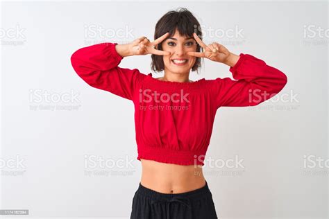 고립 된 흰색 배경 위에 서있는 빨간 여름 티셔츠를 입은 젊은 아름다운 여성은 얼굴에 손가락으로 평화 의 상징을하고 승리를