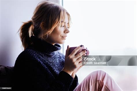 파란 스웨터를 입은 사려 깊은 소녀가 커피 한 잔을 들고 창문 에 앉아 거리를 바라봅니다 20 24세에 대한 스톡 사진 및 기타 이미지 20 24세 가정 생활 가정의