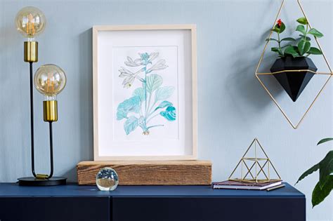 꽃 포스터와 수직 나무 프레임 테이블 램프 골드 피라미드 액세서리 회색 벽 바탕에 기하학적 냄비에 거는 식물을 모의 인테리어